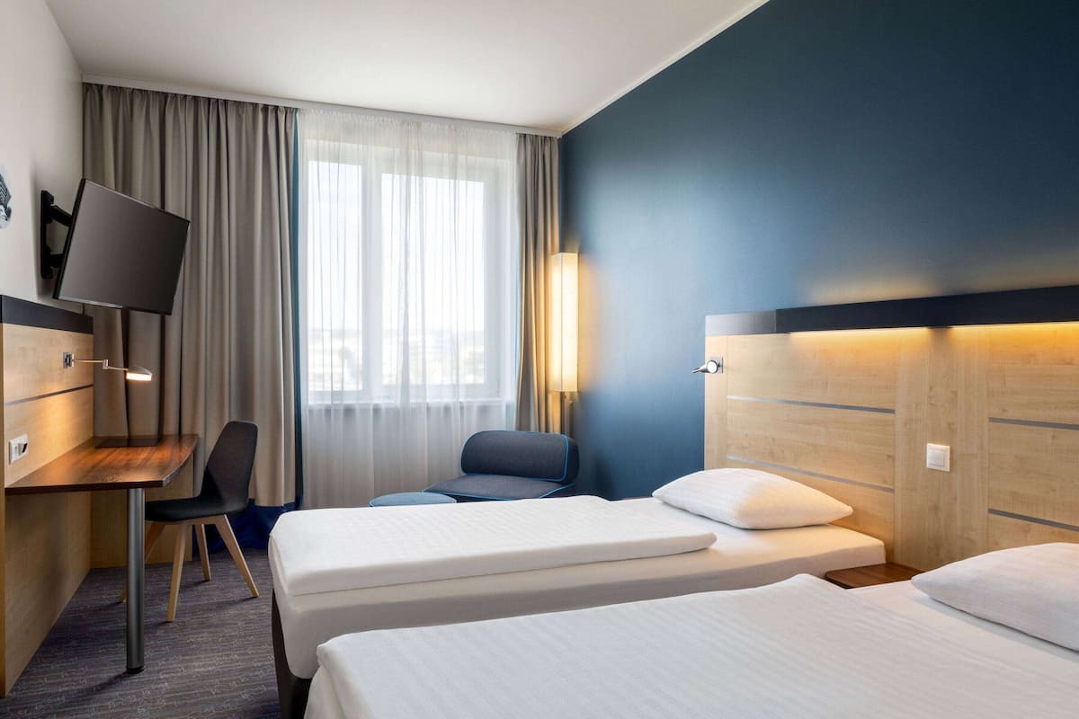 Quarto de hotel com duas camas, uma escrivaninha, uma cadeira, televisão e uma janela grande com cortinas.