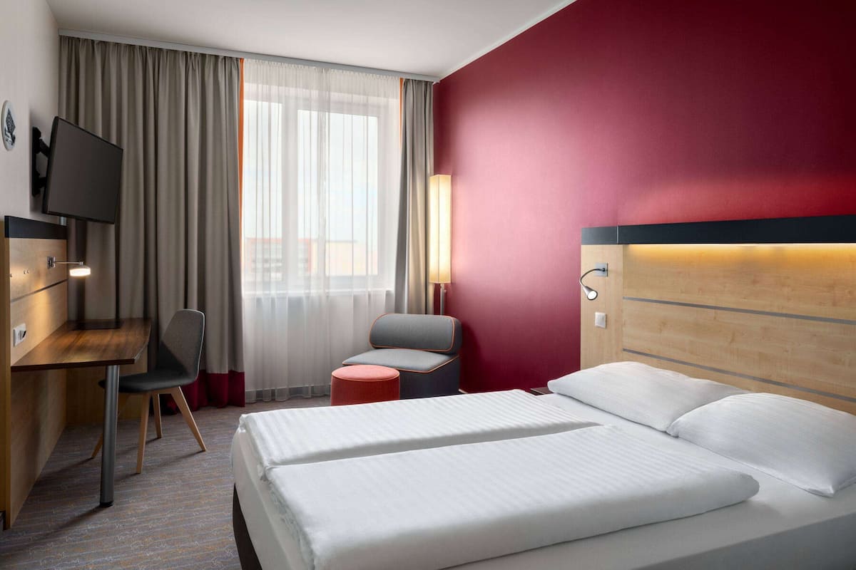 Quarto de hotel com duas camas, uma escrivaninha com cadeira, televisão e janela com cortinas.