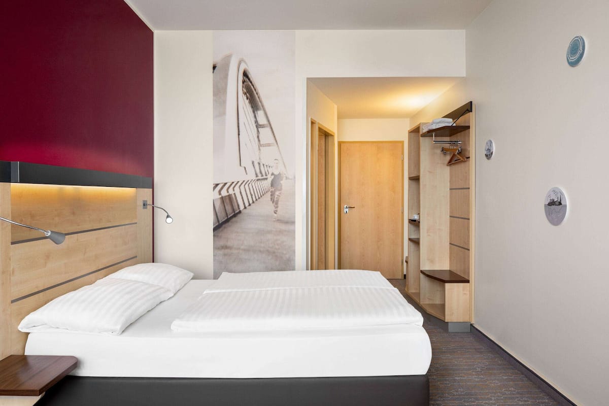 Quarto de hotel com uma cama, cabeceira de madeira, prateleira fixada na parede, guarda-roupa e uma porta que dá para outro quarto.