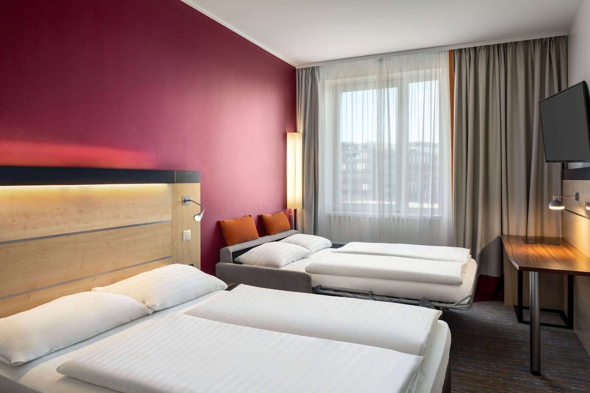 Quarto de hotel com uma cama grande, uma cama menor, uma escrivaninha e uma televisão.