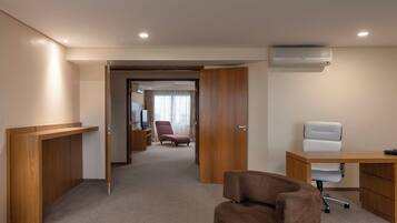 Quarto de hotel com escrivaninha de madeira, uma cadeira marrom, uma cadeira de escritório branca, televisão e ar condicionado.