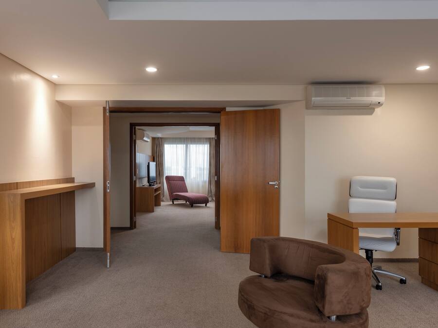 Quarto de hotel com escrivaninha de madeira, uma cadeira marrom, uma cadeira de escritório branca, televisão e ar condicionado.