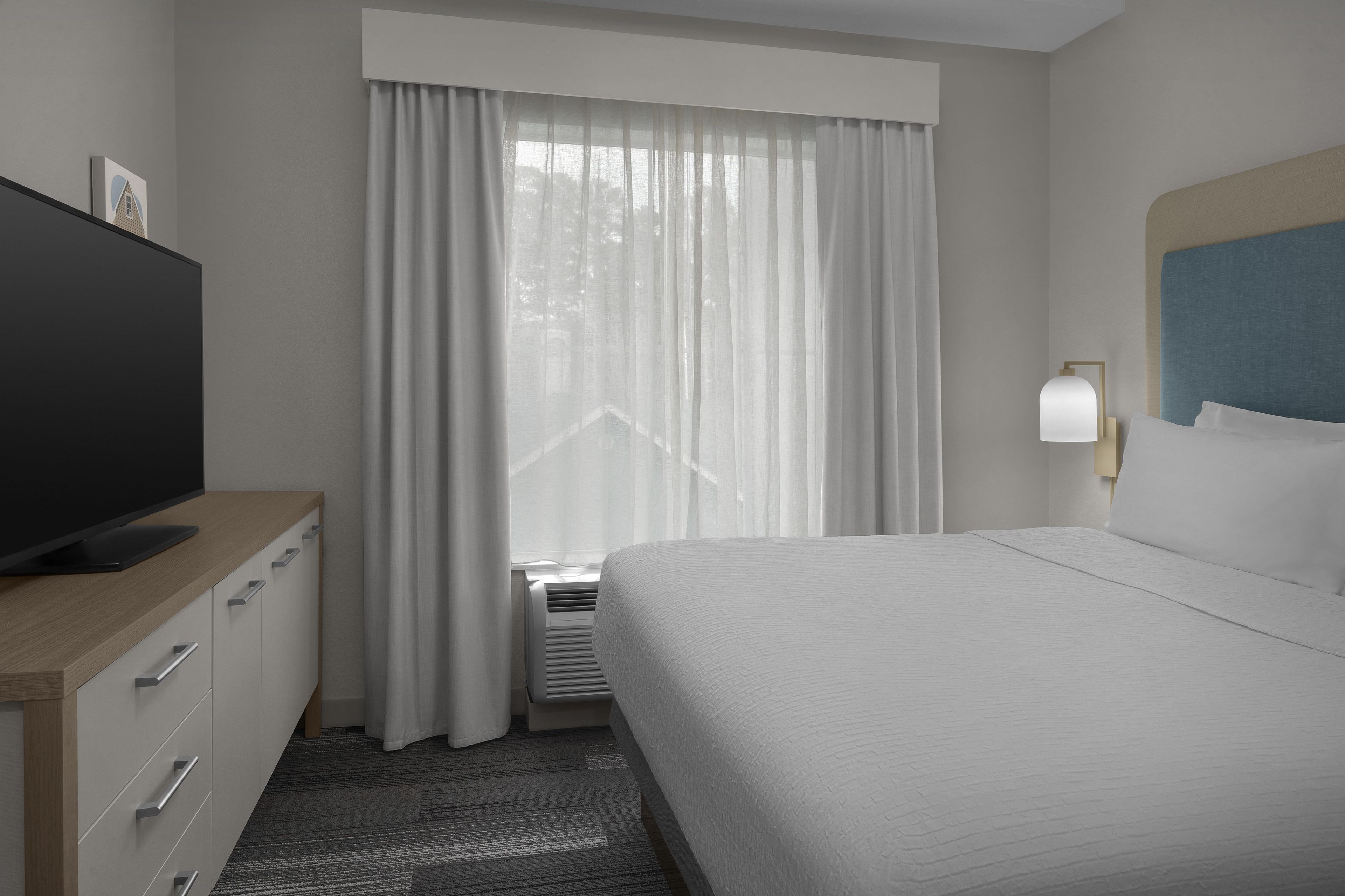Quarto de hotel com cama, televisão sobre uma cômoda de madeira, janela com cortinas leves e luminária fixada na parede.