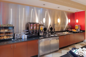 Cozinha de hotel com estação de café da manhã em autosserviço, incluindo uma máquina de waffles, cafeteira e diversos alimentos.