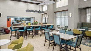 Um hall de hotel moderno com bar central, diversas opções de assento incluindo mesas e cadeiras, e grandes janelas que proporcionam luz natural.