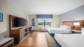 Quarto de hotel com uma televisão grande, uma escrivaninha, uma cadeira, uma cama, uma mesa de cabeceira e uma janela com vista para o exterior.