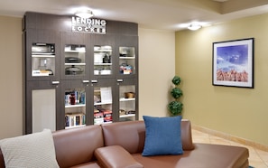 Quarto com sofá de couro marrom, um armário 'Lending Locker' com diversos itens, uma moldura na parede e uma pequena planta.