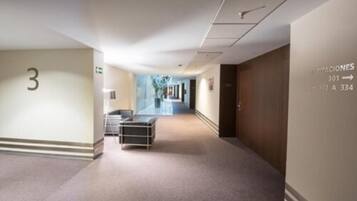 Corredor de hotel com piso de carpete, um banco e uma parede com números de quartos e uma placa de sinalização.
