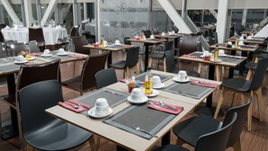 Um restaurante com mesas postas para refeições, cadeiras e vista da cidade através de grandes janelas.