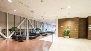 Um hall amplo com uma receção em madeira, sofás pretos e plantas em vasos.