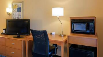 Quarto de hotel com escrivaninha, televisão, micro-ondas, cadeira e um pequeno frigorífico.
