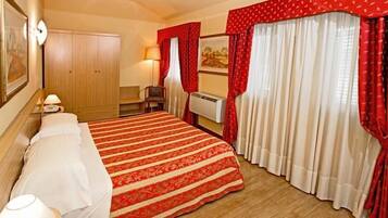 Quarto de hotel com uma cama grande, um guarda-roupa de madeira, uma secretária com cadeira e uma janela com cortinas vermelhas.