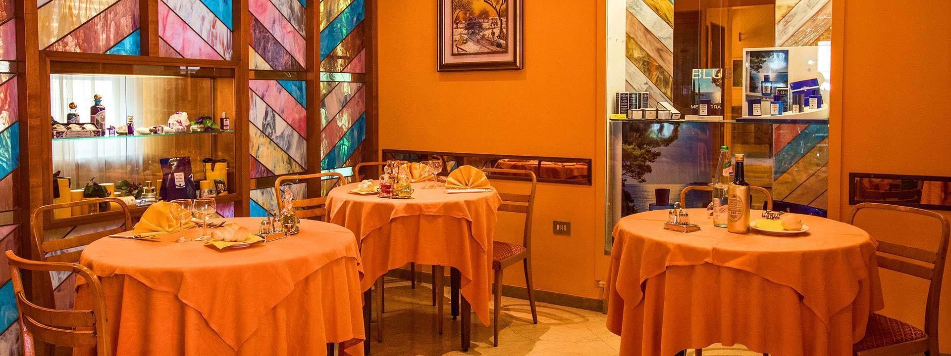 Um restaurante com três mesas redondas, cada uma coberta por uma toalha laranja. As paredes exibem padrões geométricos coloridos, e há prateleiras com garrafas e copos.