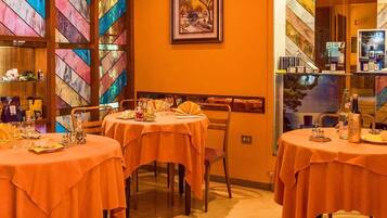 Um restaurante com três mesas redondas, cada uma coberta por uma toalha laranja. As paredes exibem padrões geométricos coloridos, e há prateleiras com garrafas e copos.