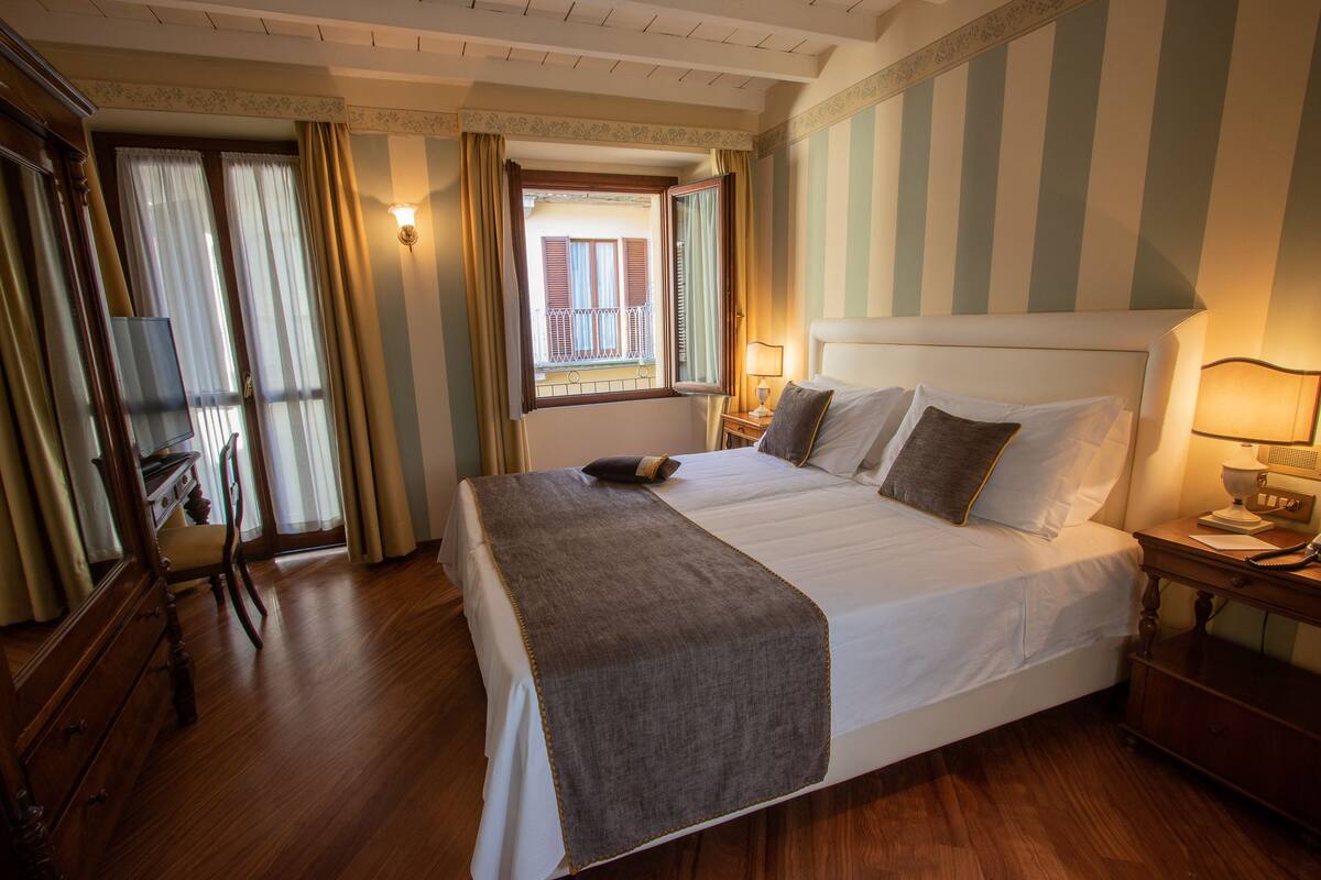 Quarto de hotel com uma cama grande, piso de madeira, cômoda, espelho e janela com cortinas.