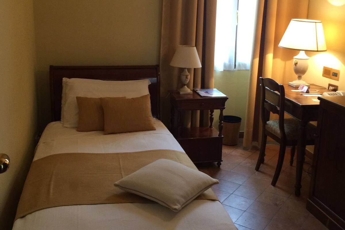 Quarto de hotel com cama, escrivaninha, cadeira, luminária e janela com cortinas.