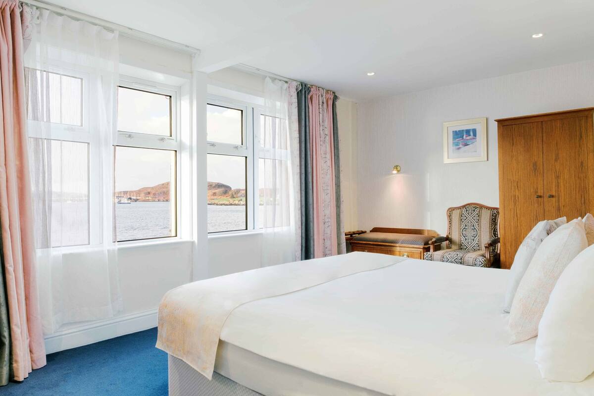 Quarto de hotel com uma cama grande, duas janelas com cortinas, um guarda-roupa de madeira, uma cadeira e uma mesa pequena.