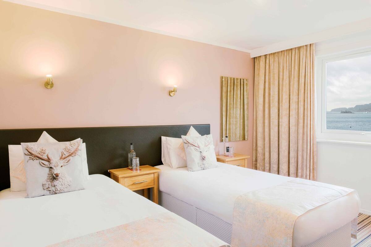Quarto de hotel com duas camas, cabeceira, mesas de cabeceira, arandelas e uma janela com cortinas, oferecendo vista para o mar.