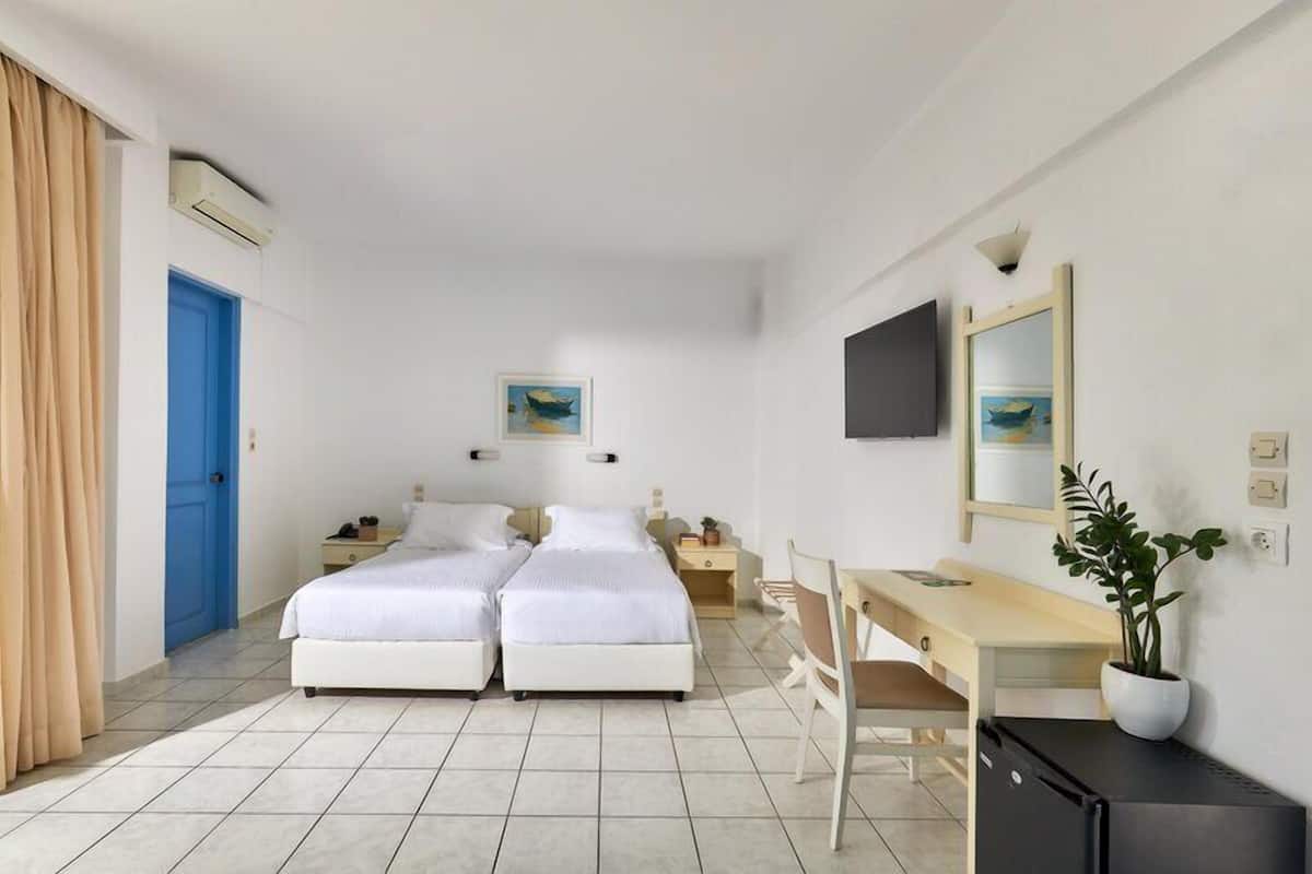 Quarto de hotel com duas camas, uma escrivaninha, uma cadeira, televisão, espelho e uma planta.