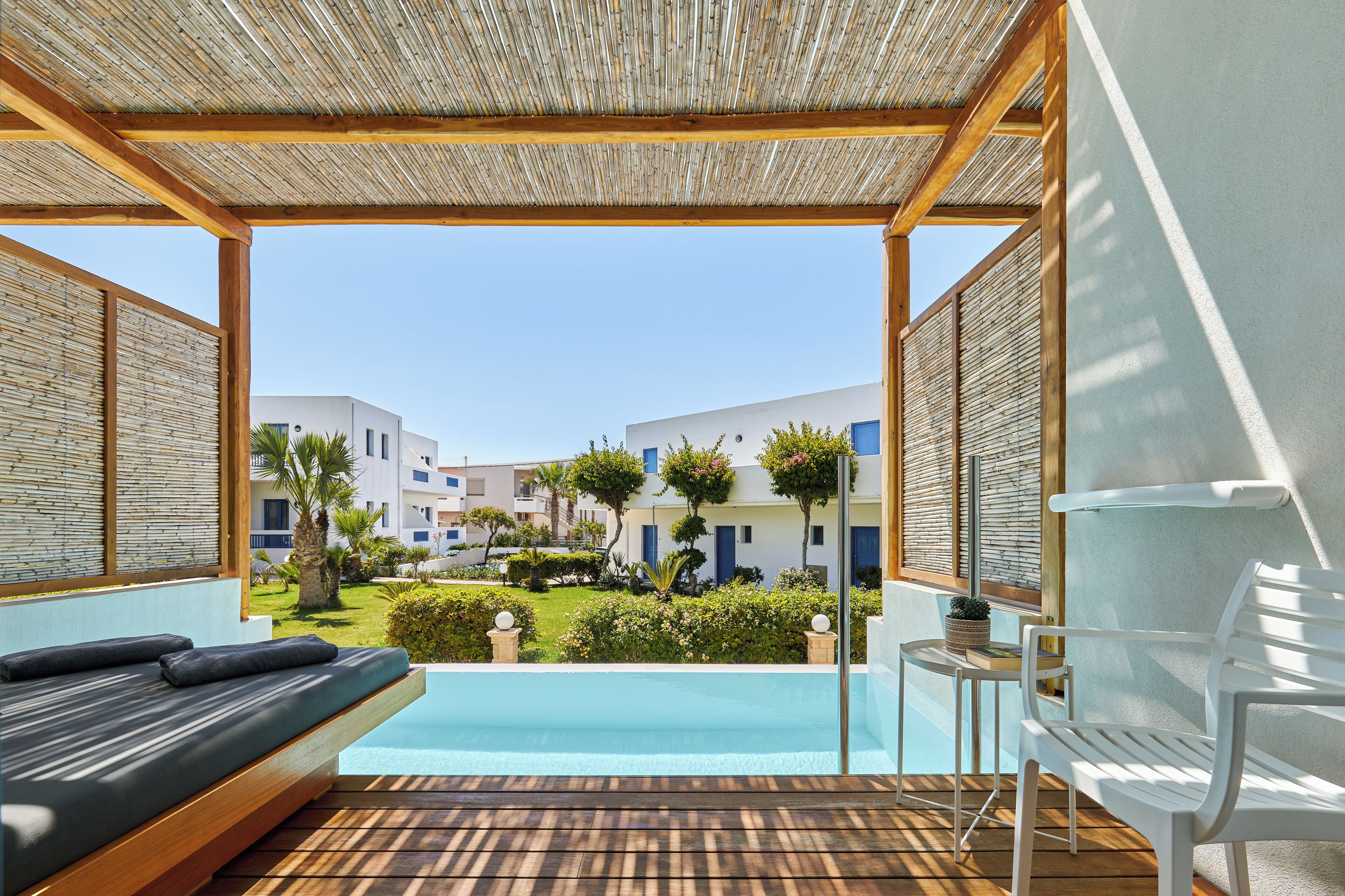 Uma varanda com piso de madeira, uma cadeira branca, uma mesinha com uma planta e vista para uma piscina e edifícios ao fundo.