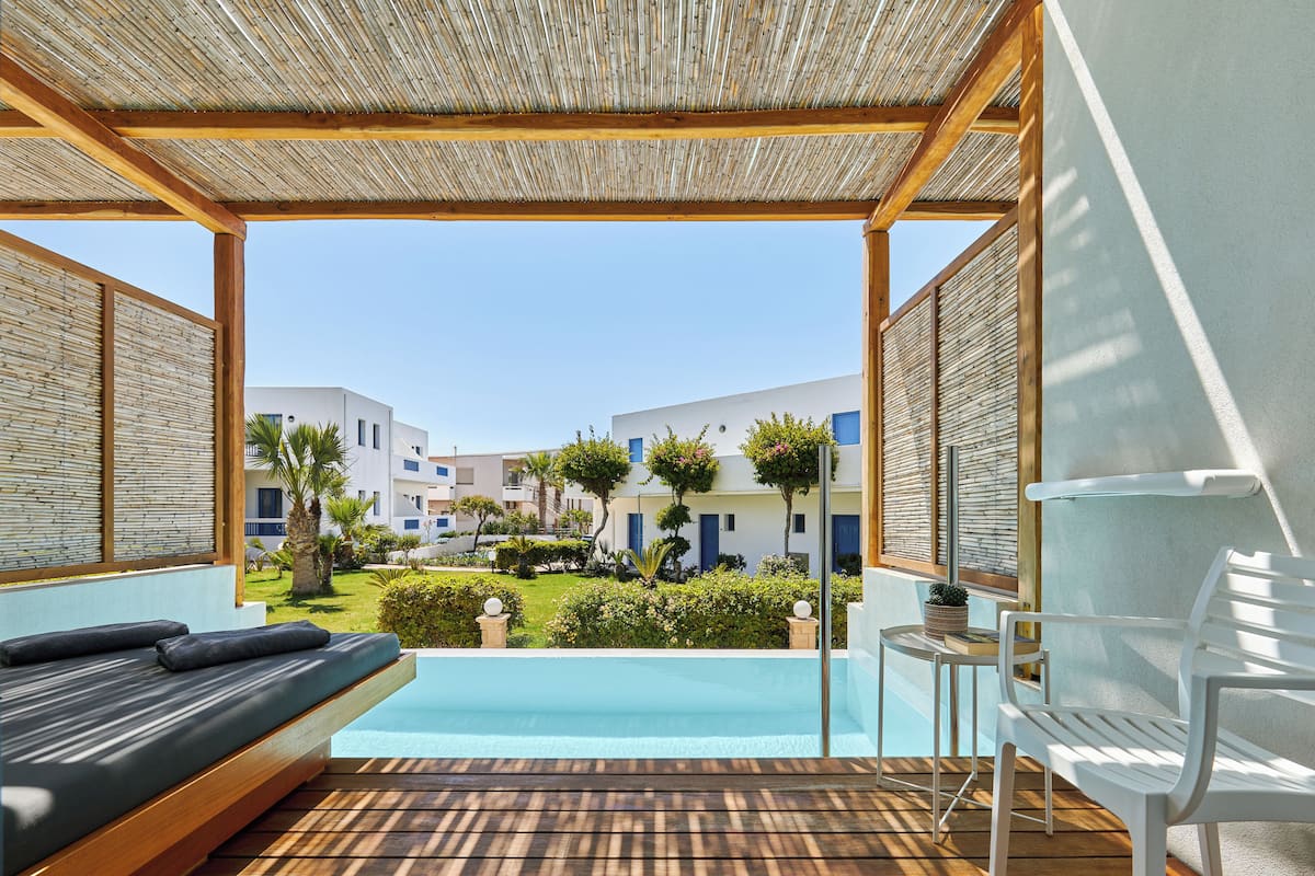 Uma varanda com piso de madeira, uma cadeira branca, uma mesinha com uma planta e vista para uma piscina e edifícios ao fundo.
