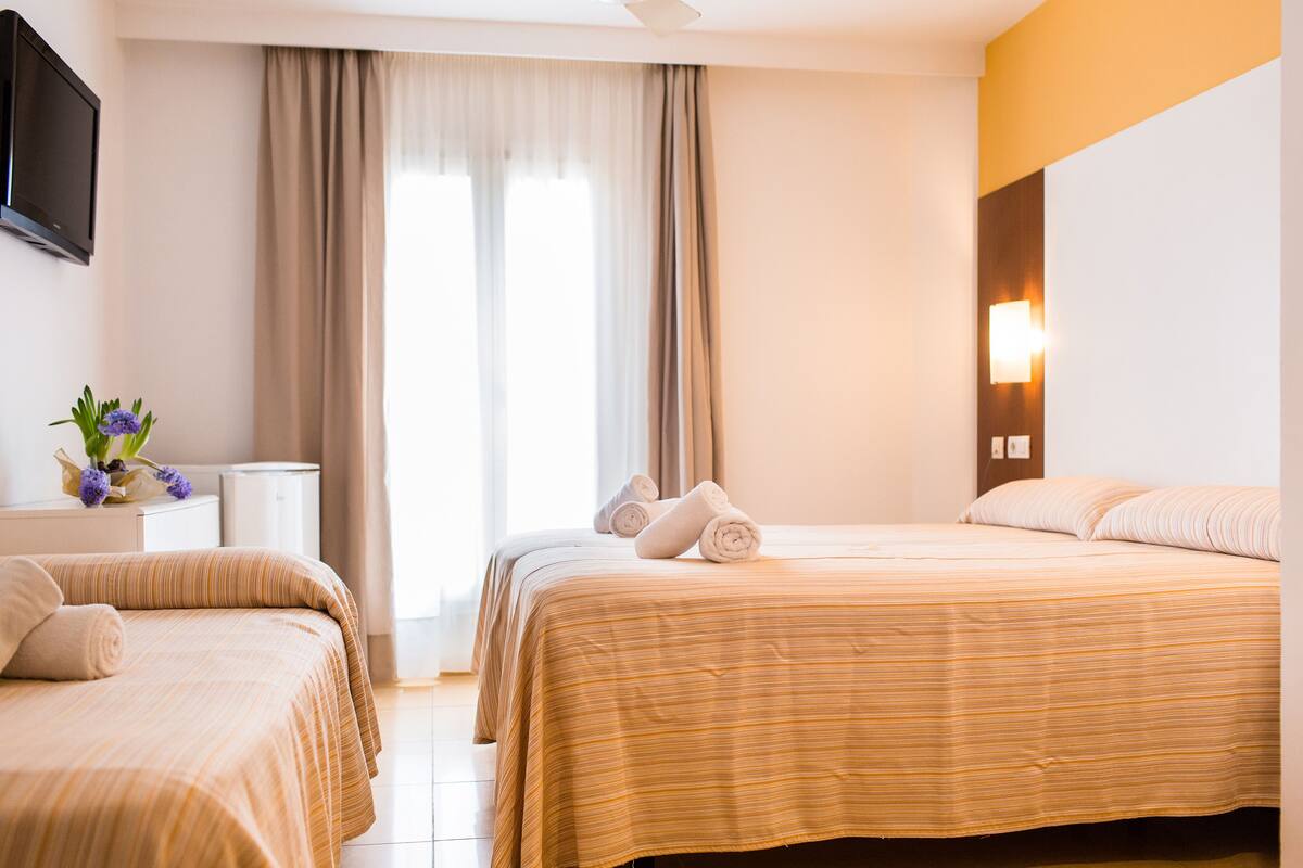 Quarto de hotel com duas camas, uma televisão de tela plana, uma mesa de cabeceira com um vaso de flores e uma janela com cortinas.