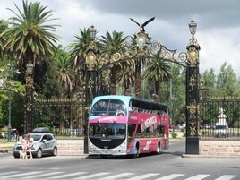 Um ônibus de dois andares com a marca 'Mendoza', passando por uma entrada com portão adornado em ferro preto e palmeiras ao fundo.