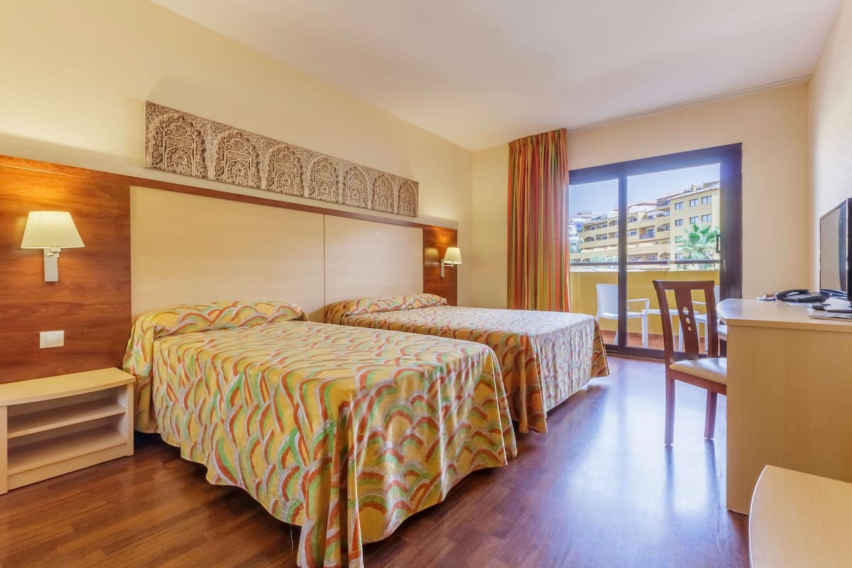 Quarto de hotel com duas camas, uma escrivaninha, uma cadeira, televisão e varanda com vista para edifícios.