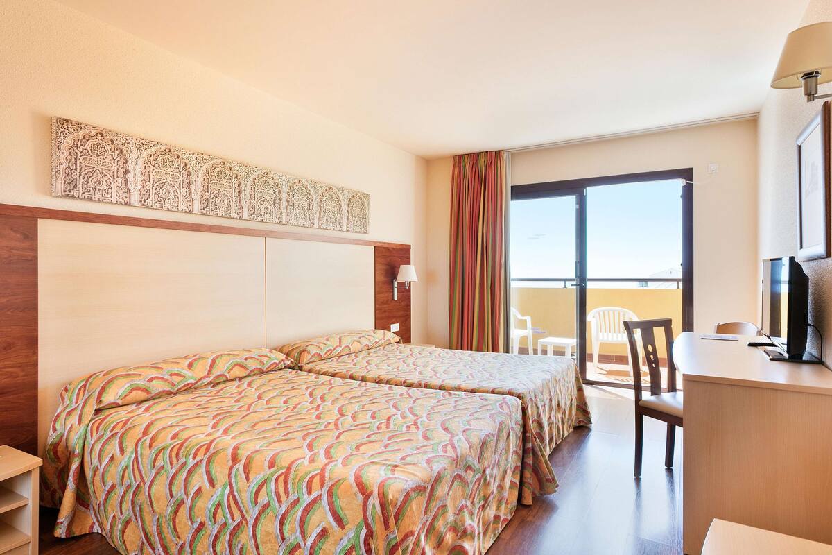 Quarto de hotel com duas camas, uma escrivaninha, uma cadeira e varanda com vista.