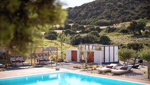 Uma área de piscina com espreguiçadeiras e um pequeno edifício, cercada por vegetação e colinas.