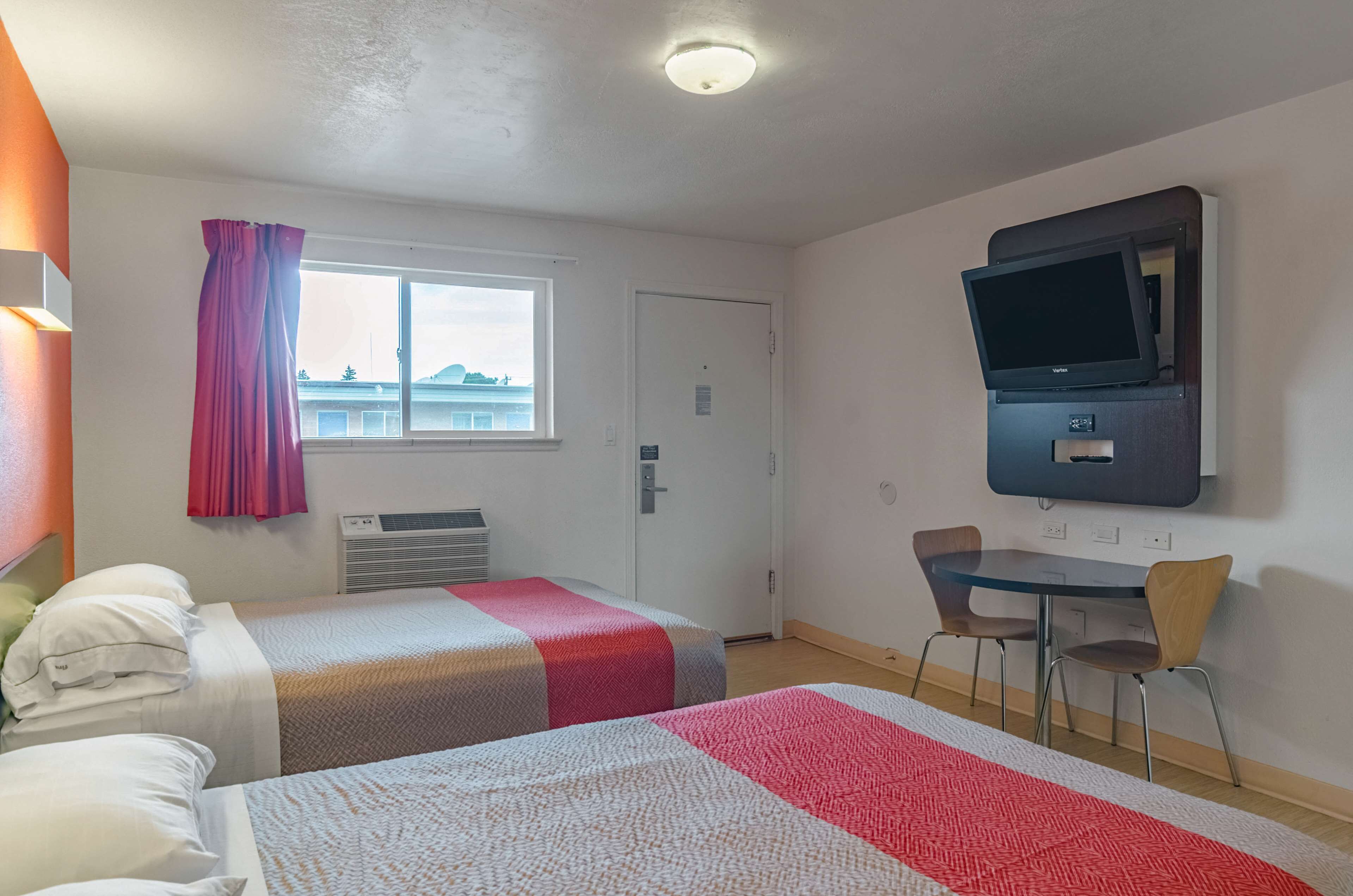 Quarto de hotel com duas camas, uma televisão presa à parede, janela com cortinas e uma pequena mesa com cadeiras.