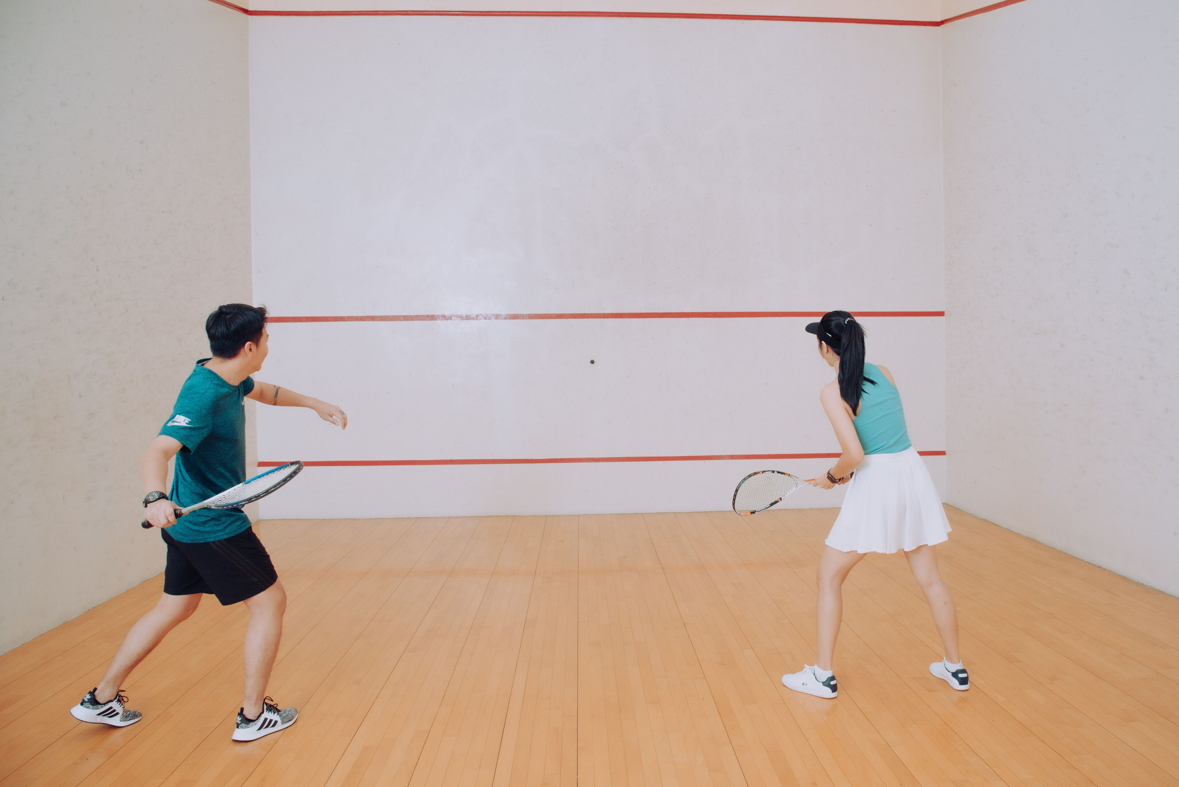 Duas pessoas jogando squash em uma sala com piso de madeira e linhas vermelhas demarcando a quadra.