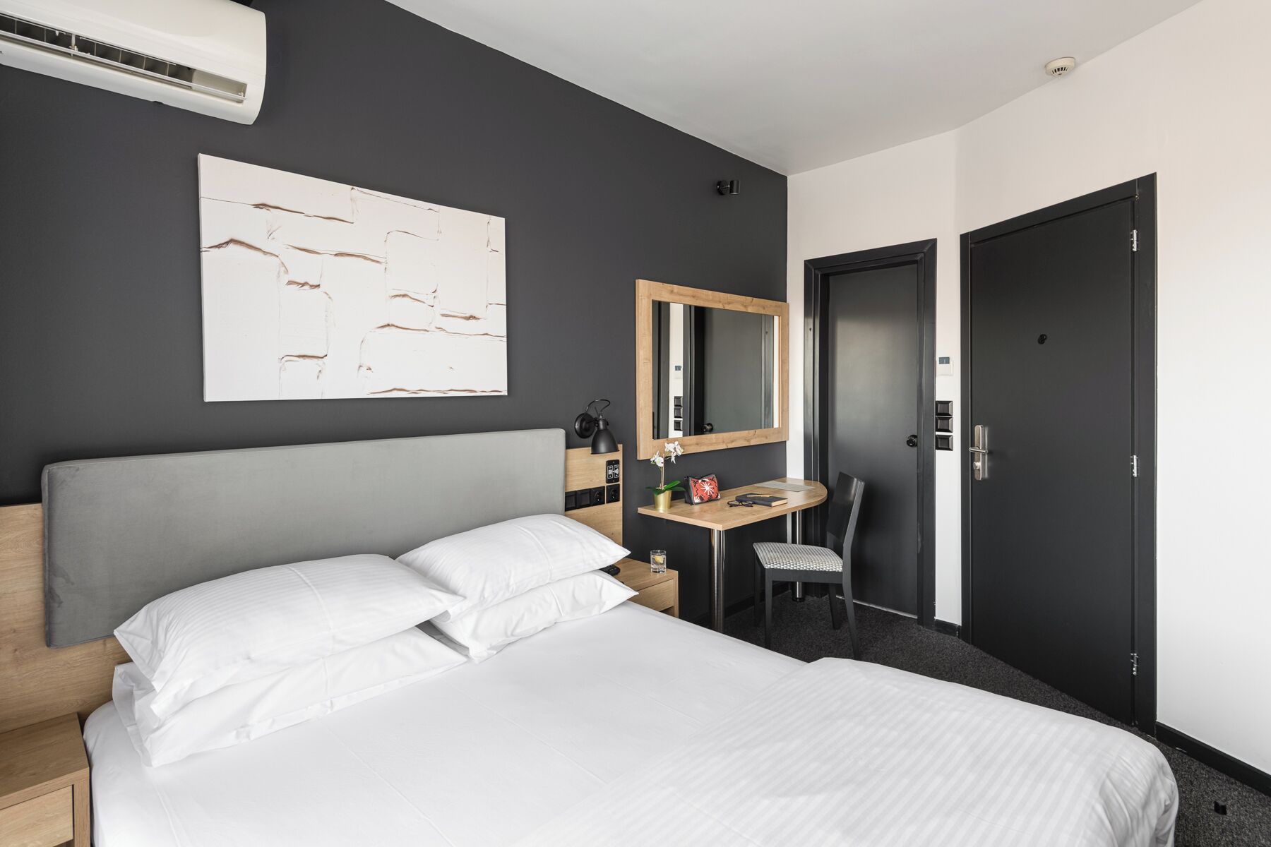 Quarto de hotel com cama, escrivaninha, cadeira, espelho e porta.