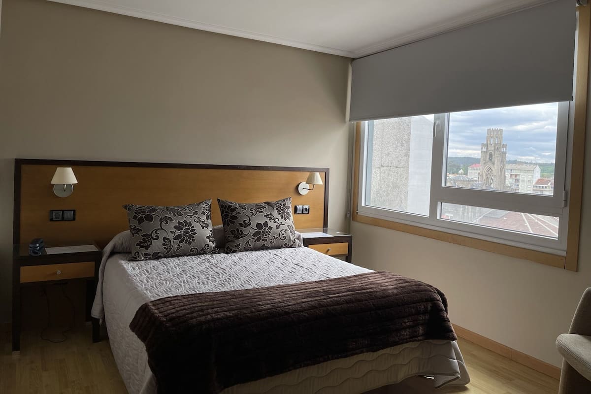 Quarto com uma cama grande, duas luminárias de cabeceira, uma cabeceira de madeira e uma janela com vista para a paisagem urbana.