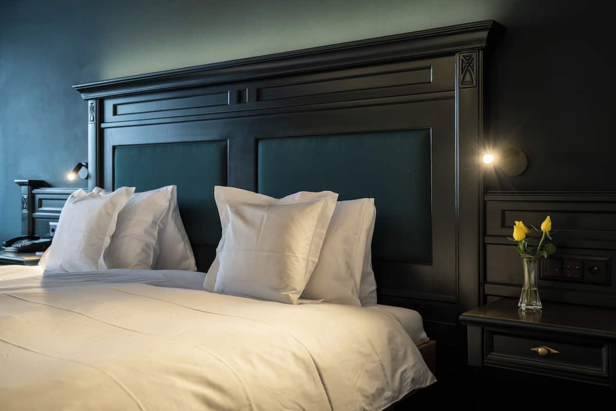 Cama bem arrumada com lençóis brancos, cabeceira escura e mesa de cabeceira com um vaso de flores amarelas.