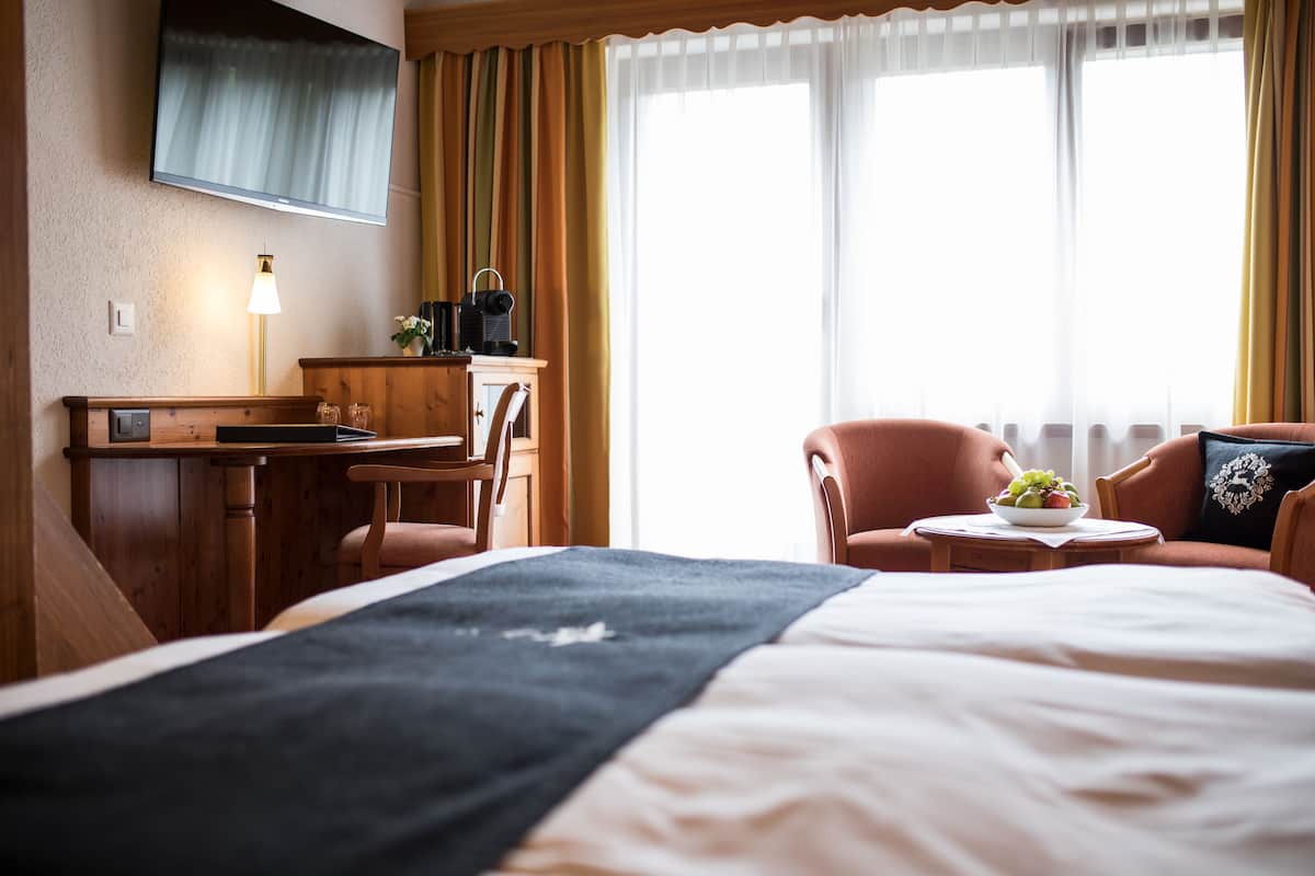 Quarto de hotel com cama, escrivaninha, cadeira, televisão fixada na parede e janela com cortinas.