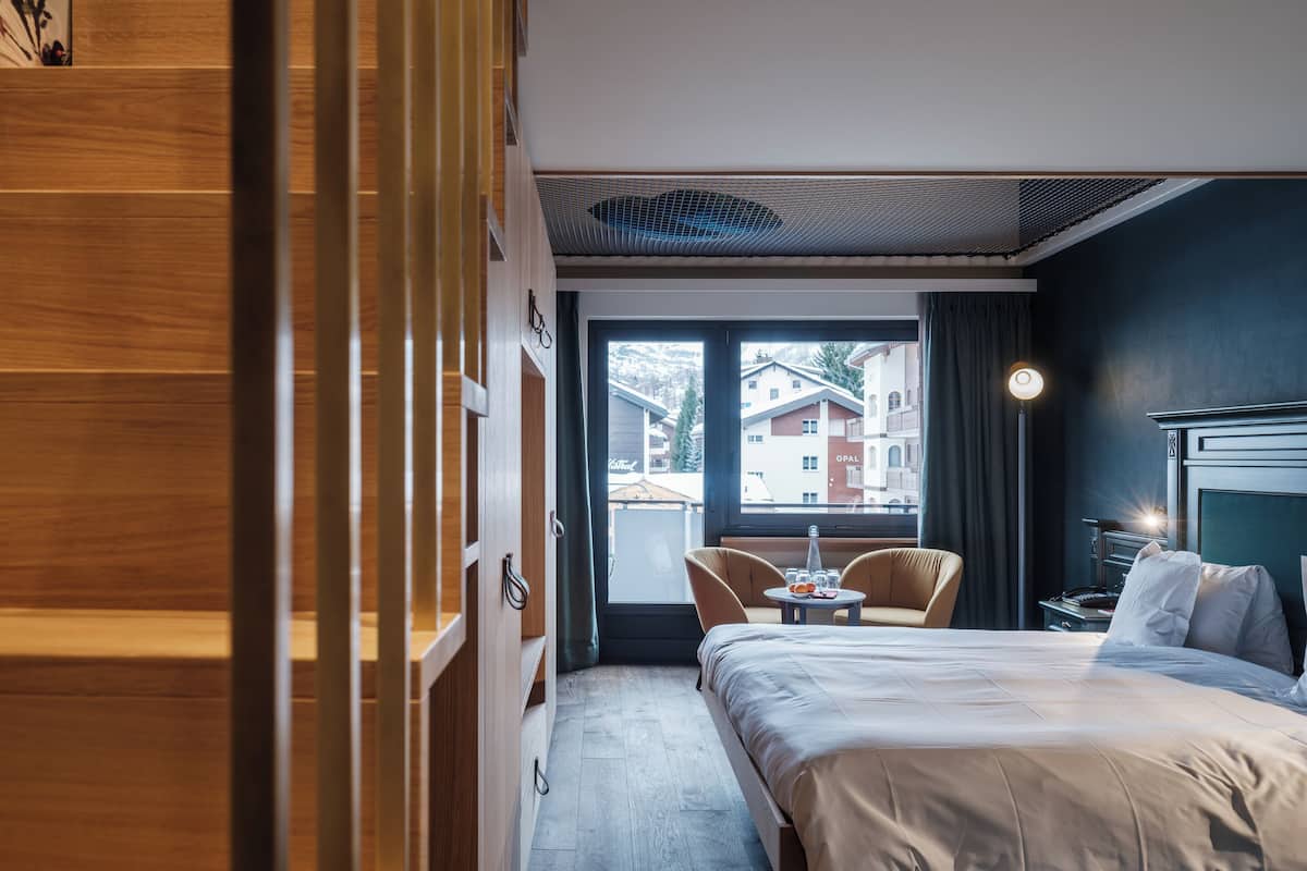 Quarto de hotel moderno com guarda-roupa de madeira, uma cama grande, um espaço de estar com uma mesa e vista para edifícios na janela.