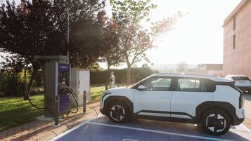 Um veículo elétrico branco carregando numa estação, com céu claro e árvores ao fundo.