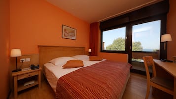 Quarto de hotel com cama, escrivaninha, cadeira, mesa de cabeceira e janela com vista.