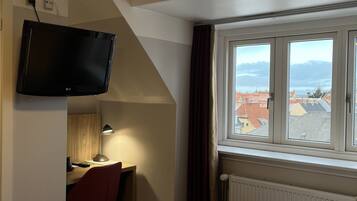 Quarto de hotel com uma cama, uma escrivaninha com cadeira, uma TV fixada na parede e uma janela com vista para edifícios e montanhas.
