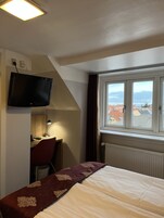 Quarto de hotel com uma cama, uma escrivaninha com cadeira, uma TV fixada na parede e uma janela com vista para edifícios e montanhas.