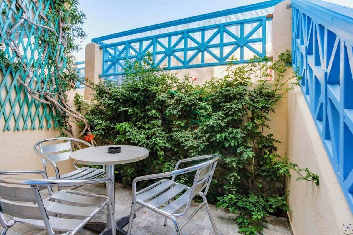 Uma pequena área externa com mesas e cadeiras, cercada por plantas e um portão de metal azul.