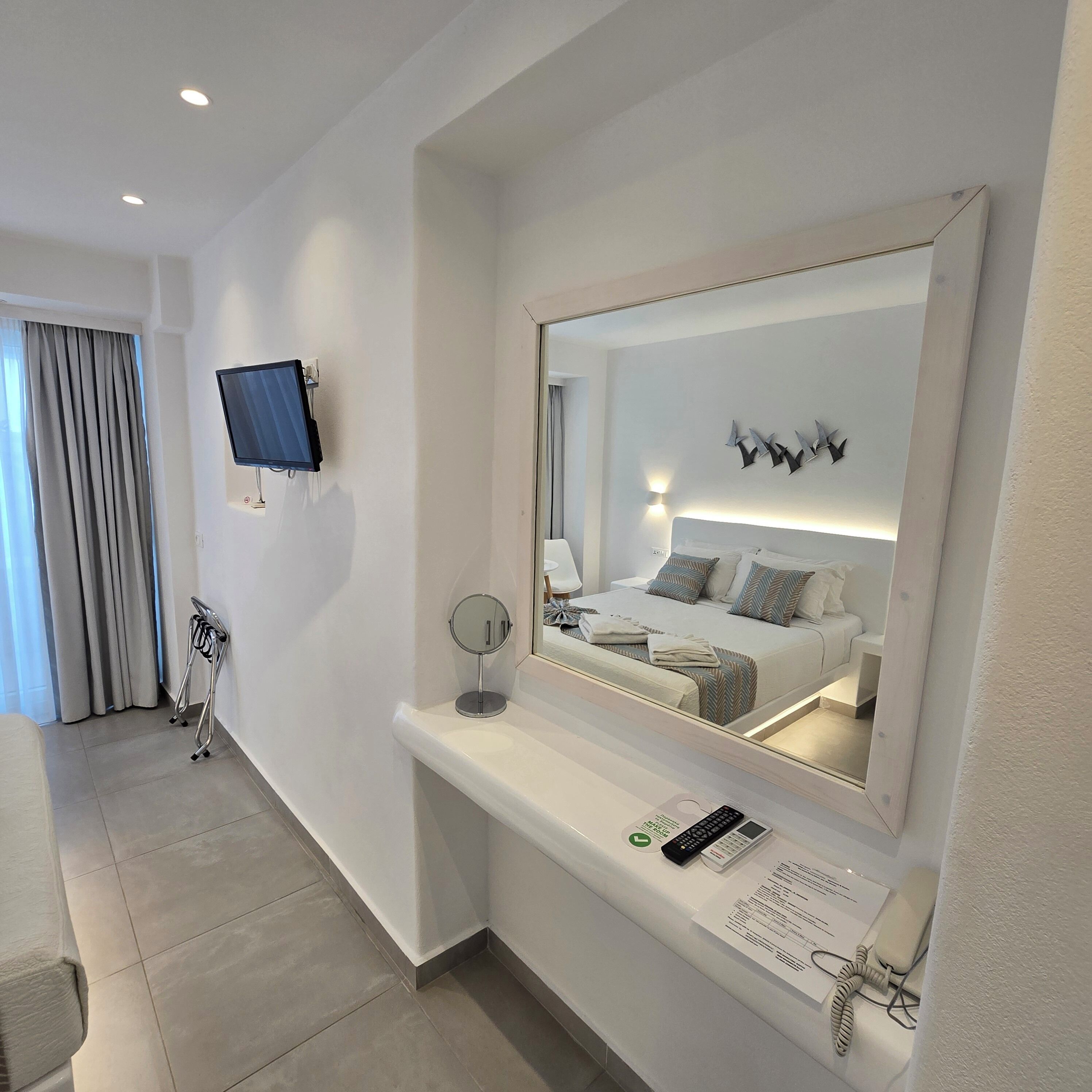 Quarto de hotel moderno com um espelho grande, mesa de cabeceira com controle remoto, telefone e TV na parede.