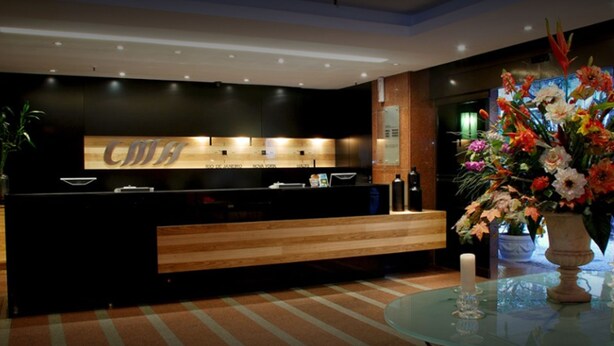 Uma área de recepção de hotel com um balcão preto, um banco de madeira, uma mesa de vidro com um vaso de flores e um arranjo floral grande.