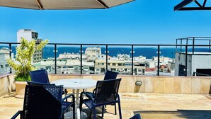 Um terraço com mesa e cadeiras, um grande guarda-sol e vista para a cidade e o mar.