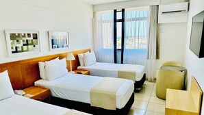 Quarto de hotel com duas camas, cabeceira de madeira, mesa de cabeceira com abajur, televisão e janela com cortinas.
