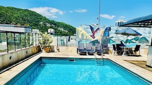 Piscina azul cristalina com espreguiçadeiras e guarda-sóis, rodeada por um deck ladrilhado e um mural de um papagaio.