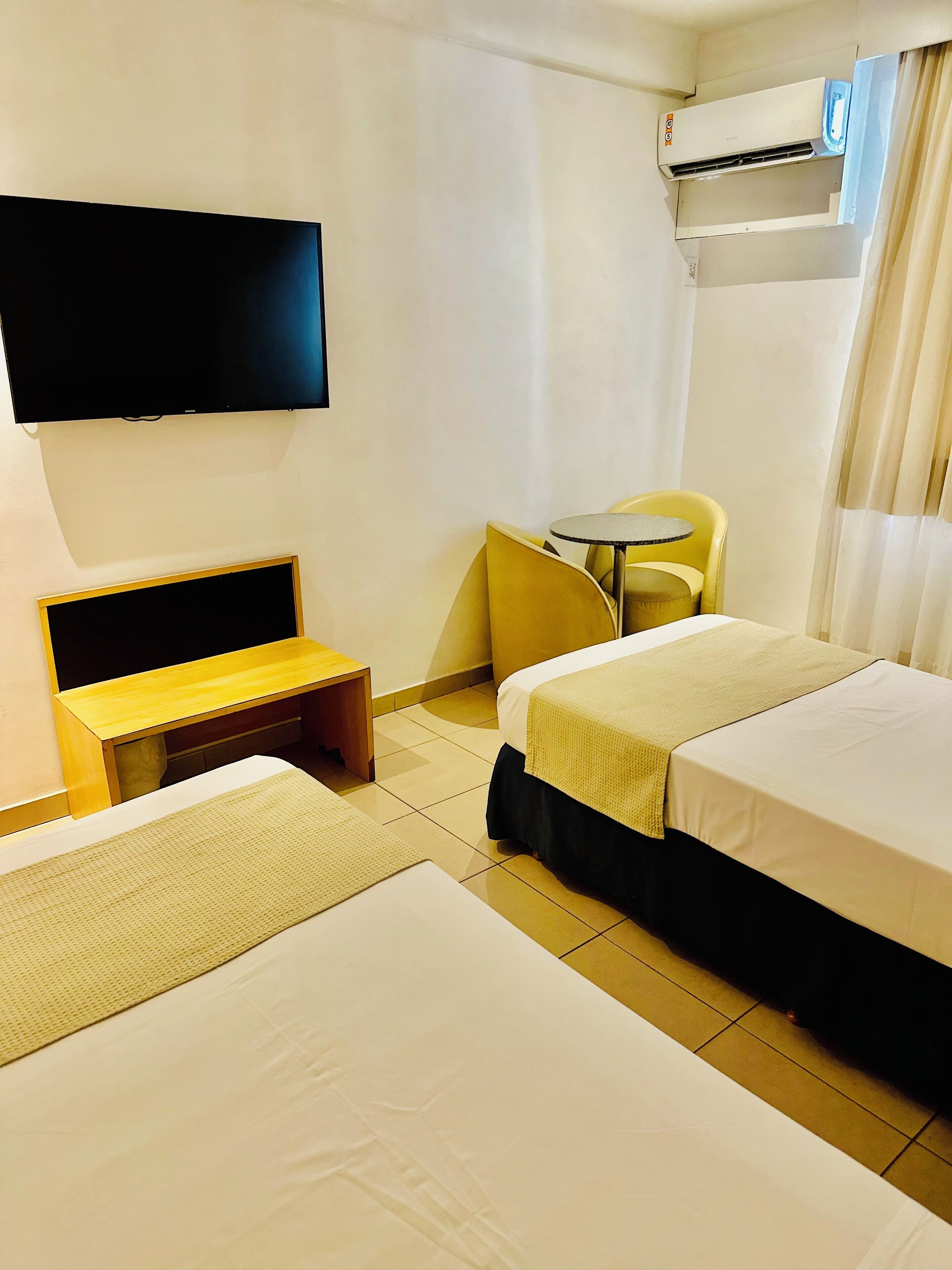 Quarto de hotel com duas camas, televisão, ar condicionado, uma mesinha e uma cadeira.