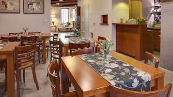 Uma área de refeições com mesas e cadeiras de madeira, um caminho de mesa floral no centro e uma bancada de cozinha com objetos decorativos.