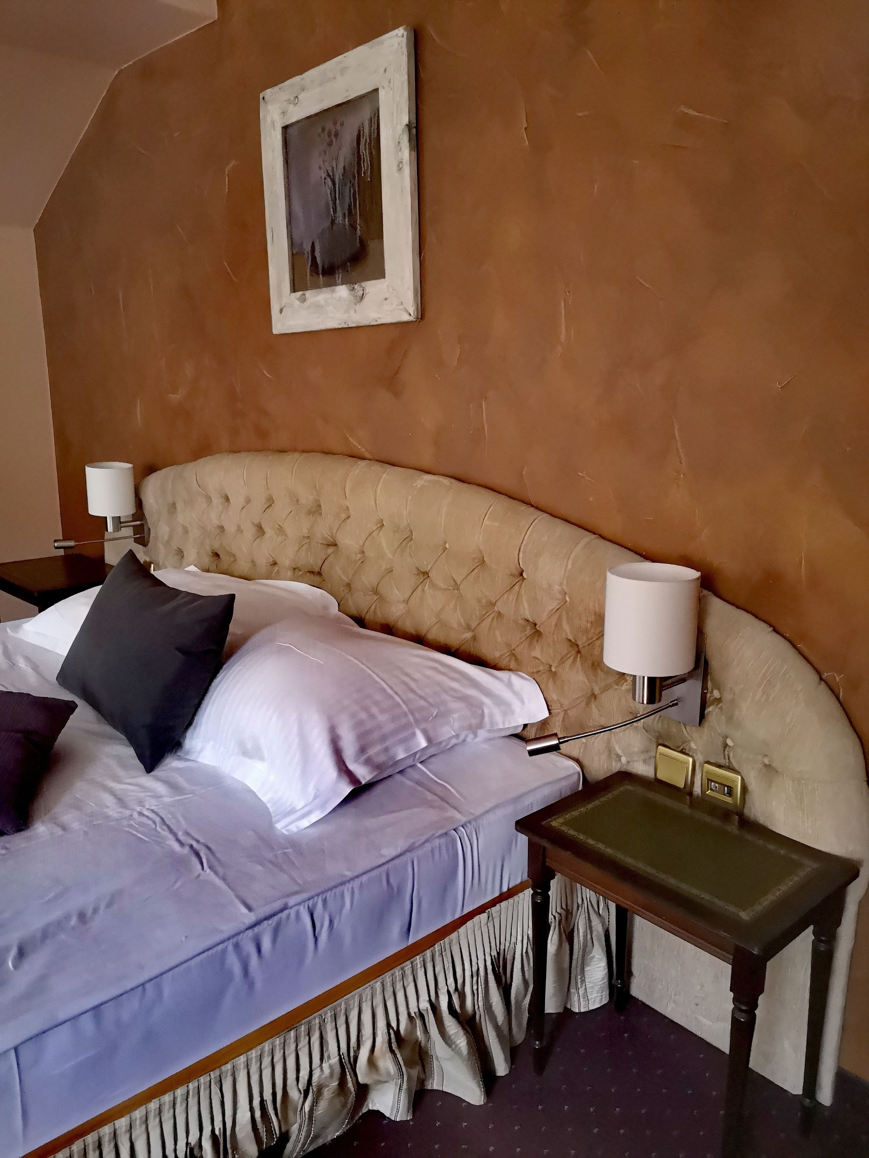 Uma cama com cabeceira estofada, várias almofadas, uma mesa de cabeceira com abajur e uma tomada.